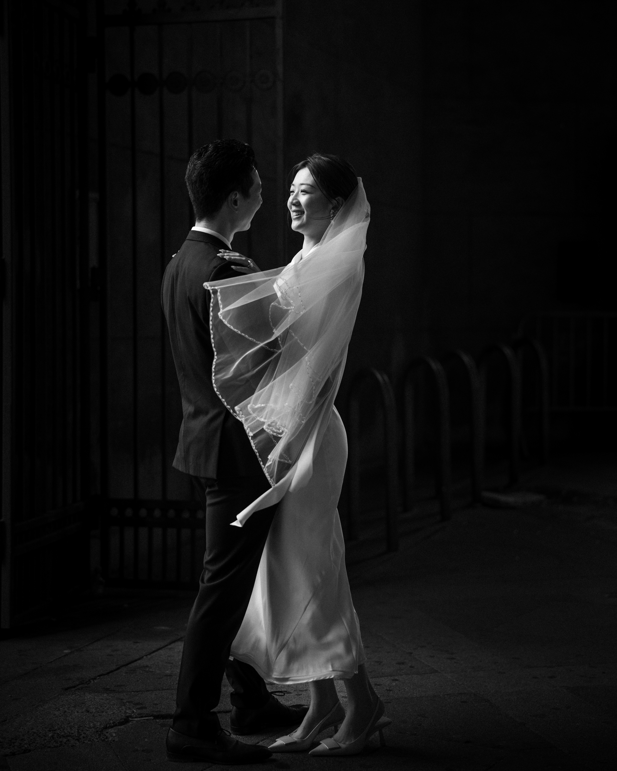 A bride and groom stand close together, smiling, as the bride’s veil ...